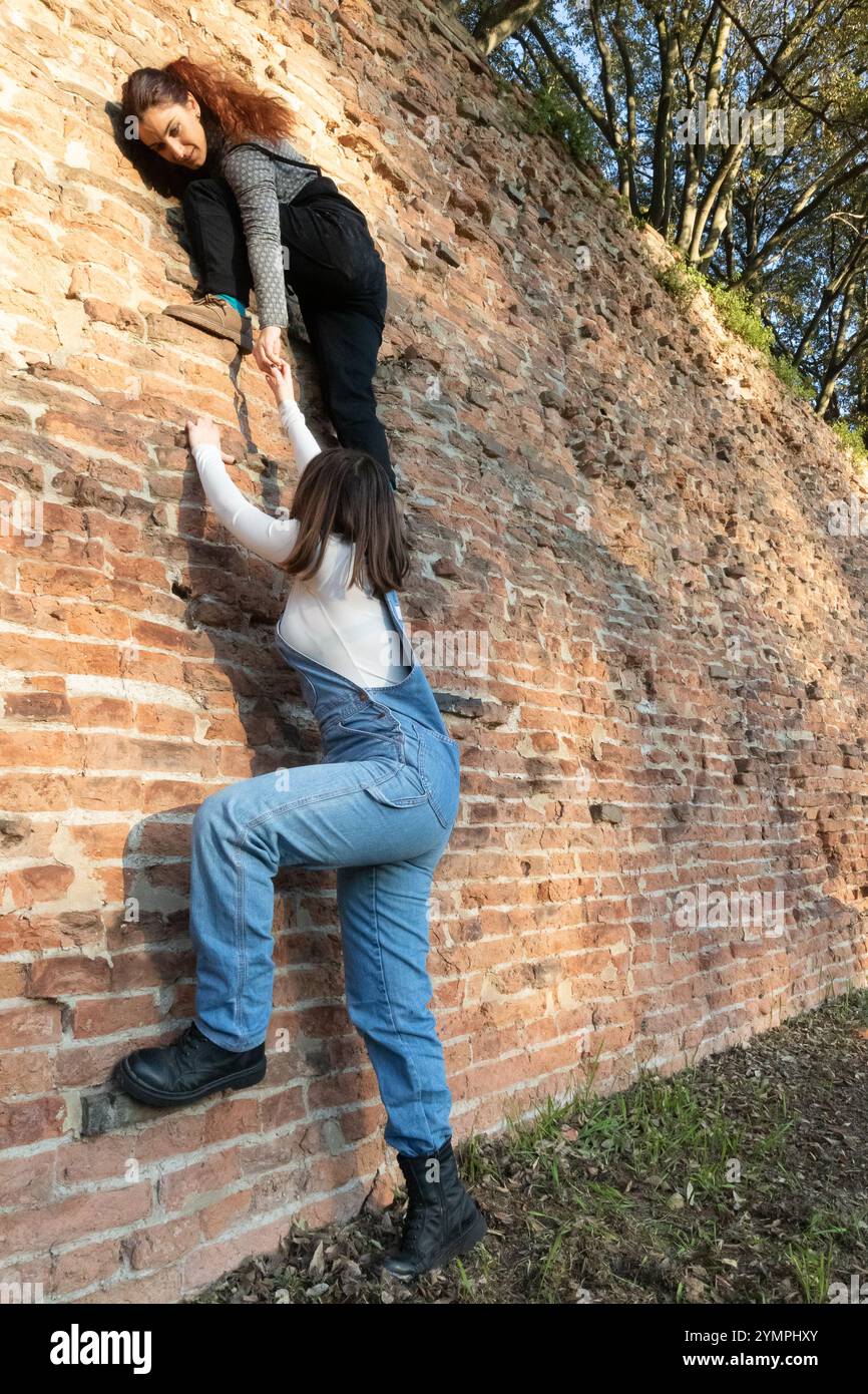 Two women climbing a brick wall, one helping the other, showcasing ...