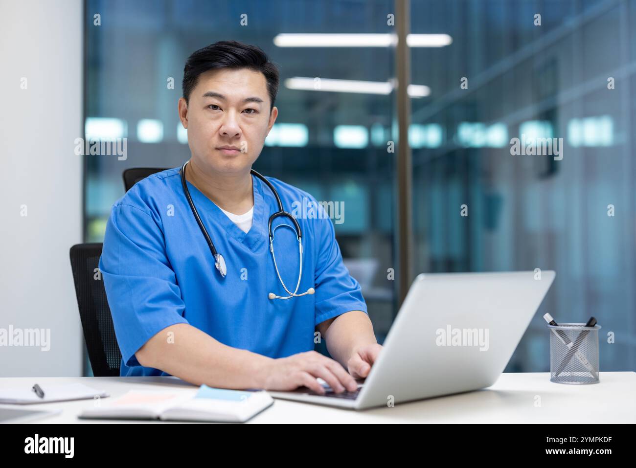 Asian man doctor wearing blue scrubs, using laptop in medical office ...