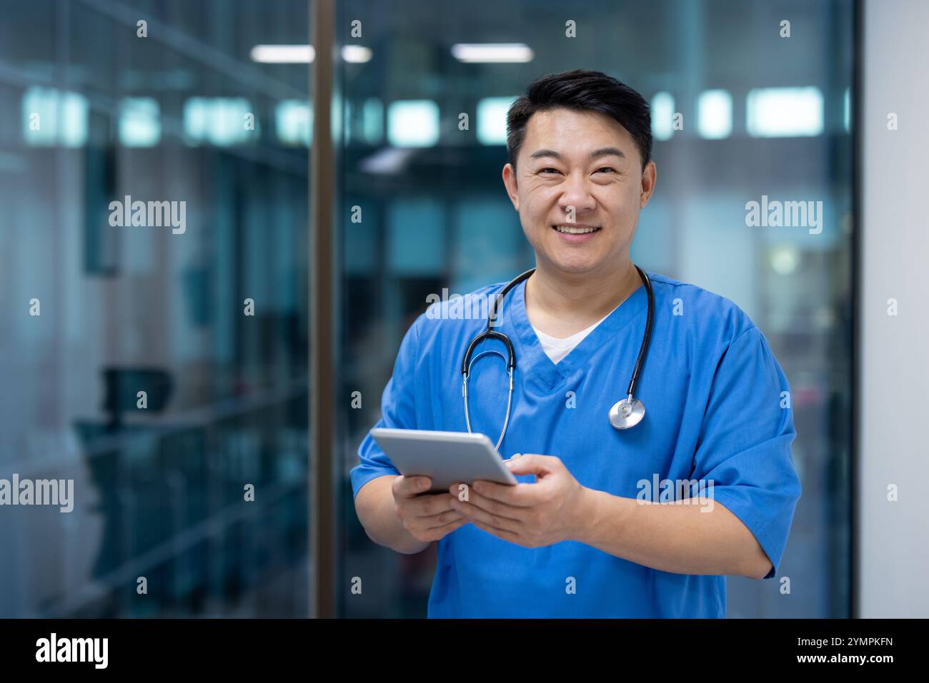 Asian man doctor wearing blue scrubs smiles while holding tablet in ...
