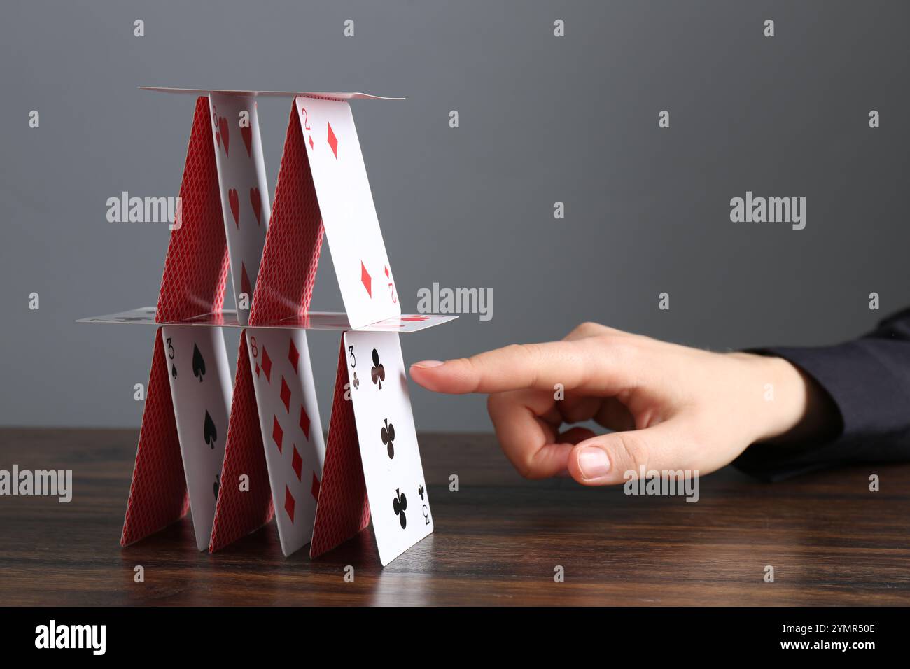 Woman destroying house of playing cards at wooden table against grey ...
