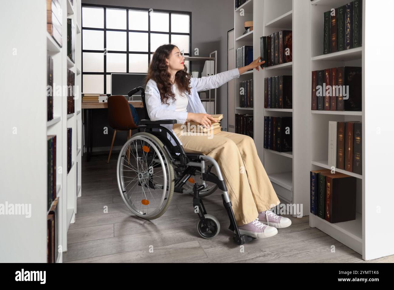 Female student in wheelchair taking book from bookshelves in library ...