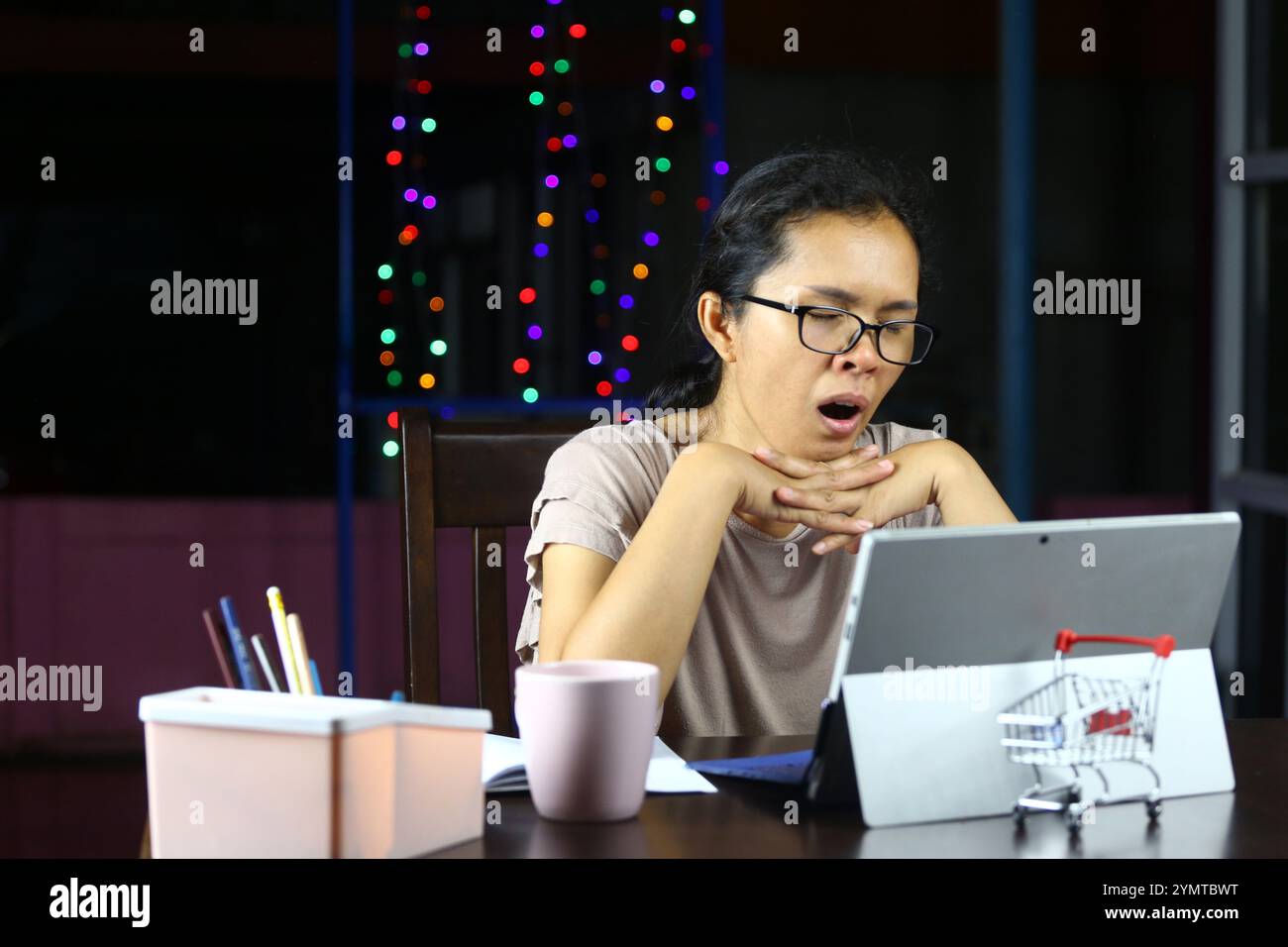 A young woman yawns while working on her tablet at home. Surrounded by ...