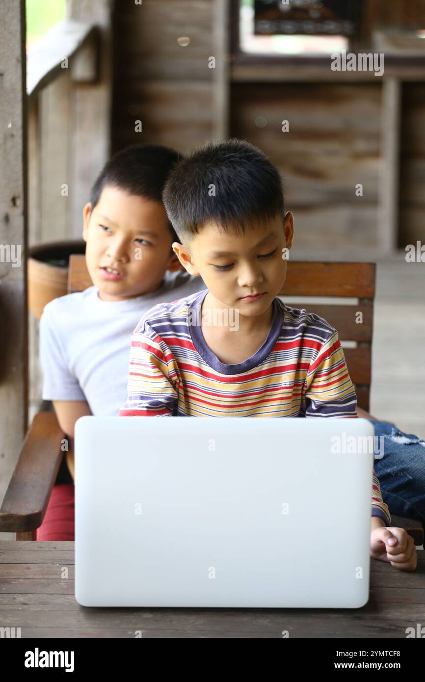 Two young boys seated in a rustic indoor setting, engaged with a laptop ...