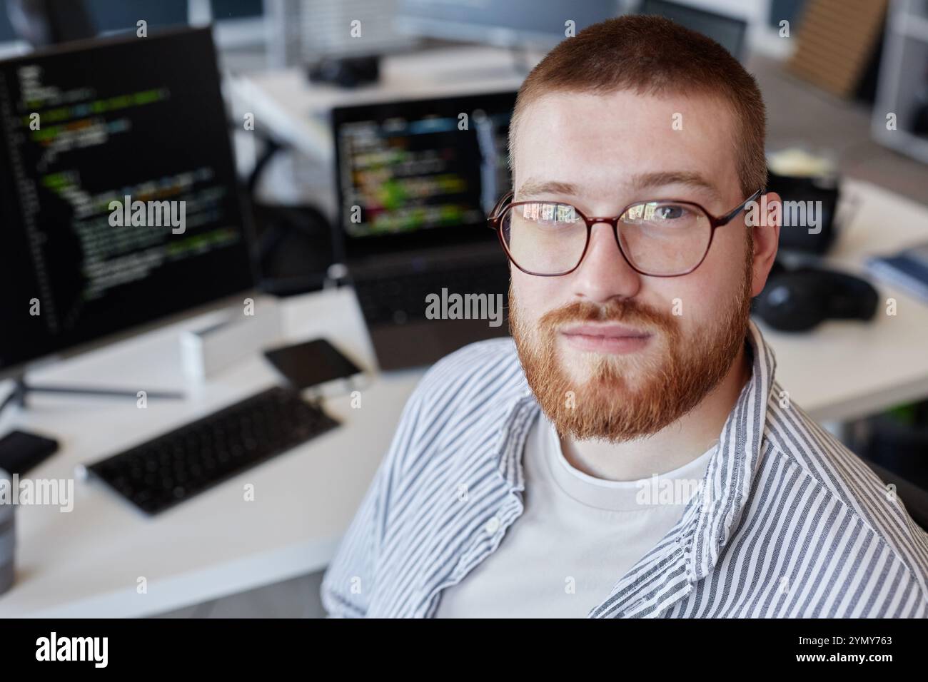Portrait of bearded man wearing glasses, seated at desk with multiple monitors showing code ...