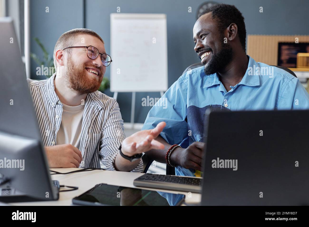 Two colleagues are sitting at a desk while smiling and discussing work ...