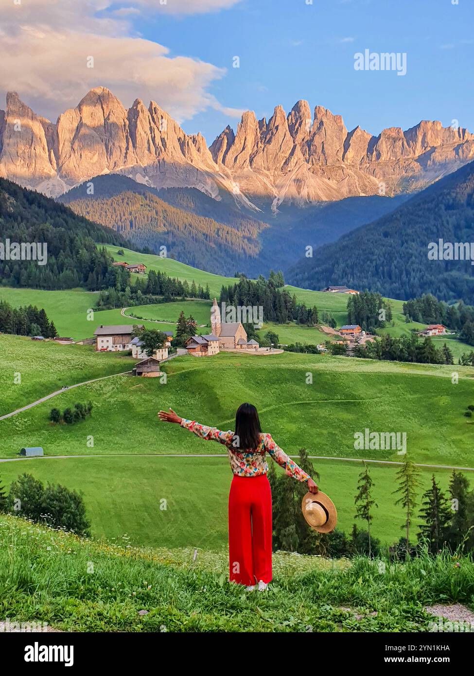 Standing atop a gentle slope, a woman in bright red pants raises her arms in joy while admiring ...