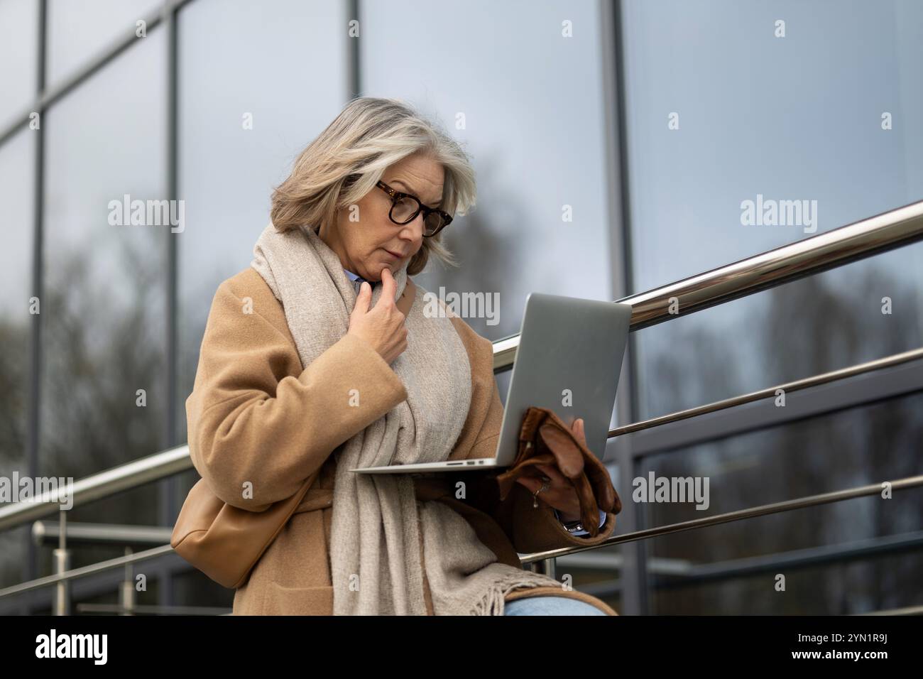 Older woman working on a laptop outdoors while seated on a railing in ...