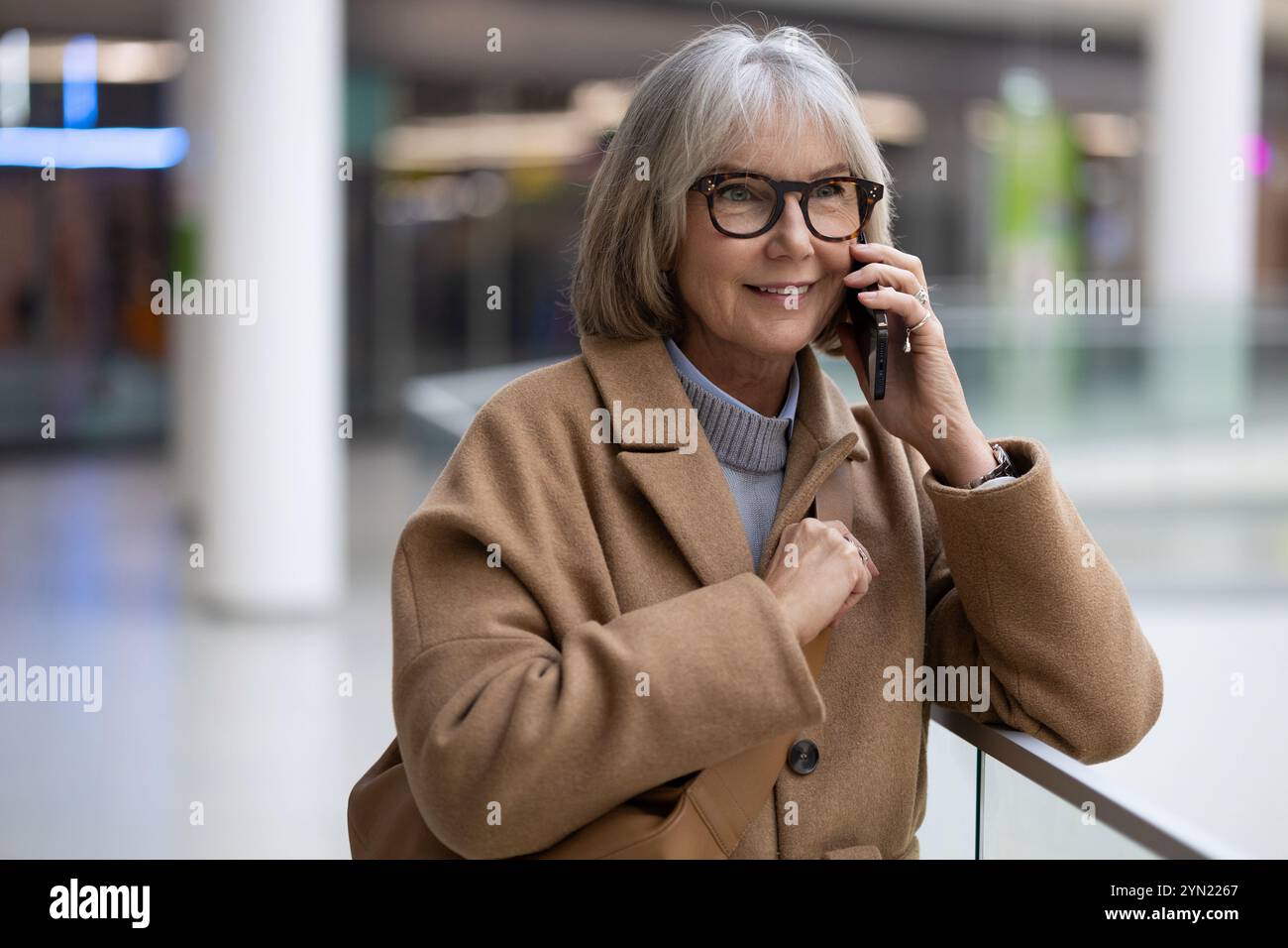 Senior woman wearing glasses engages in a phone conversation at a modern shopping center during ...
