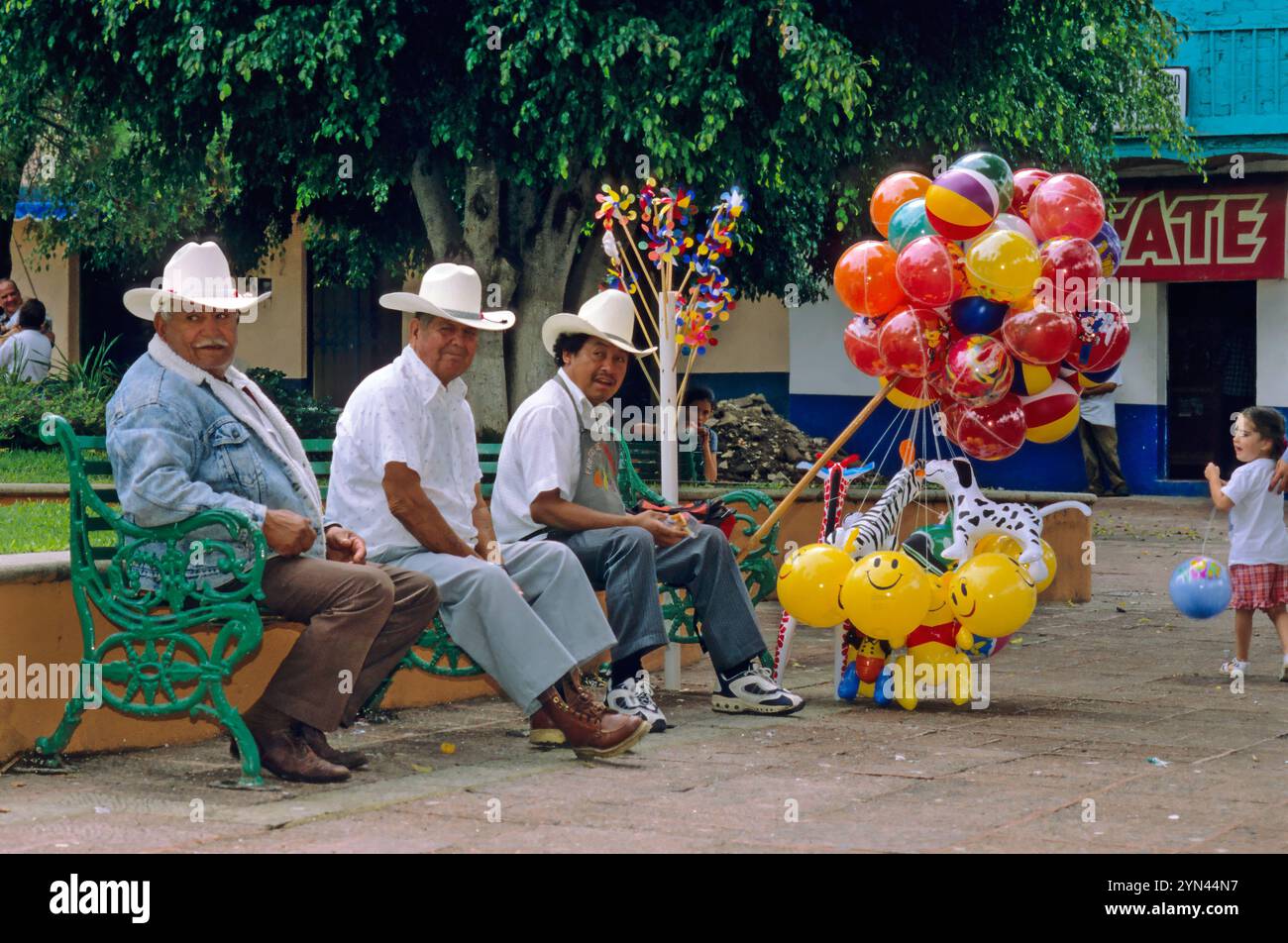 Balloon seller, men in hats at park bench at central plaza in town of ...