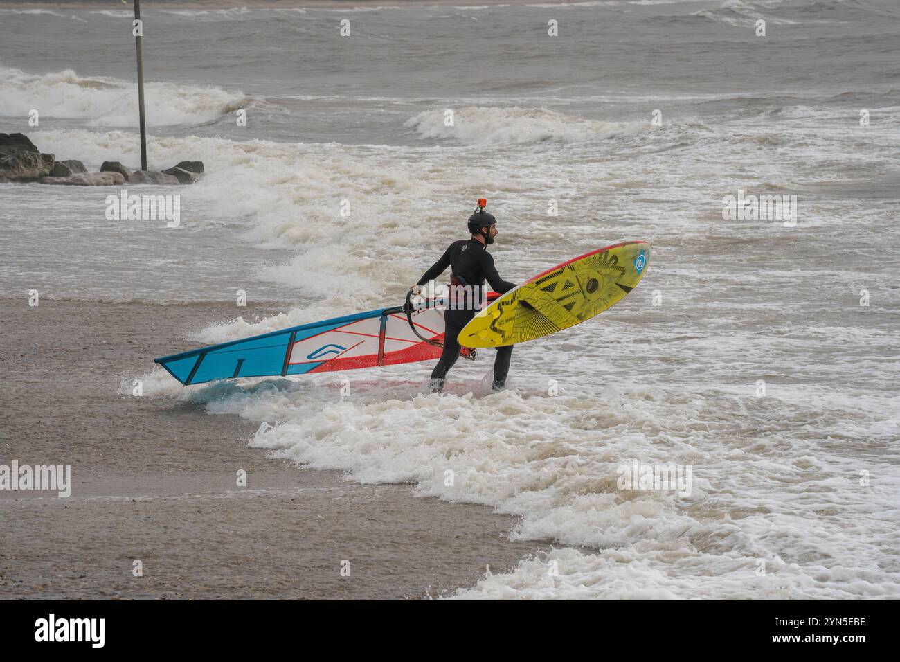 A man in a black wetsuit and helmet walks into the sea holding a ...