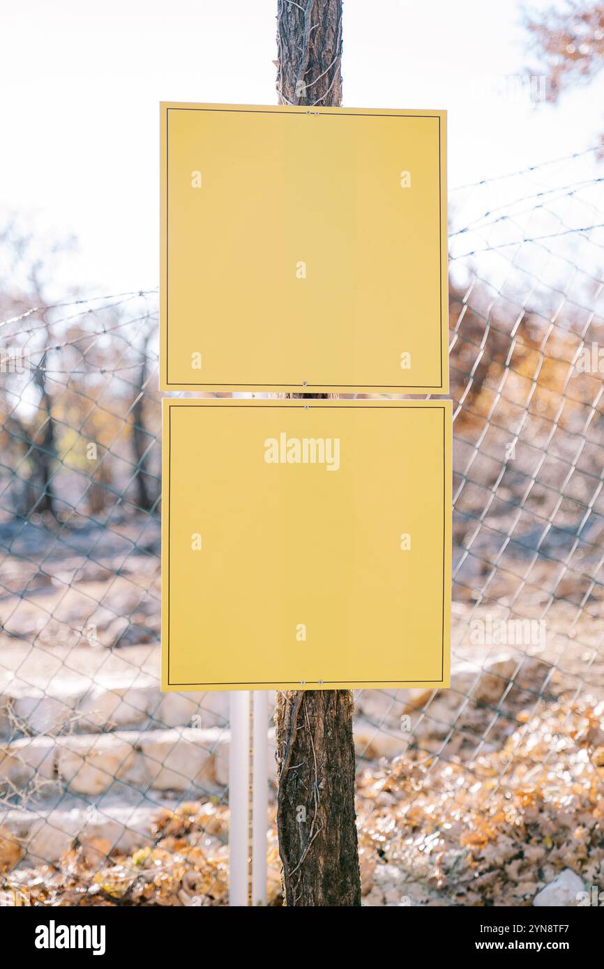 Two yellow square blank signs hang on a pole near a chain link fence ...