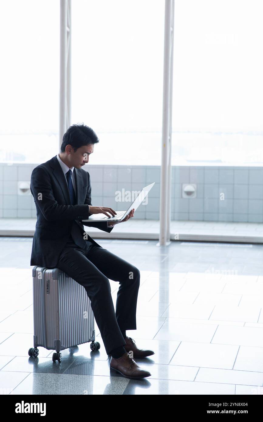 Man in his 20s in suit sitting on carrying case operating laptop ...