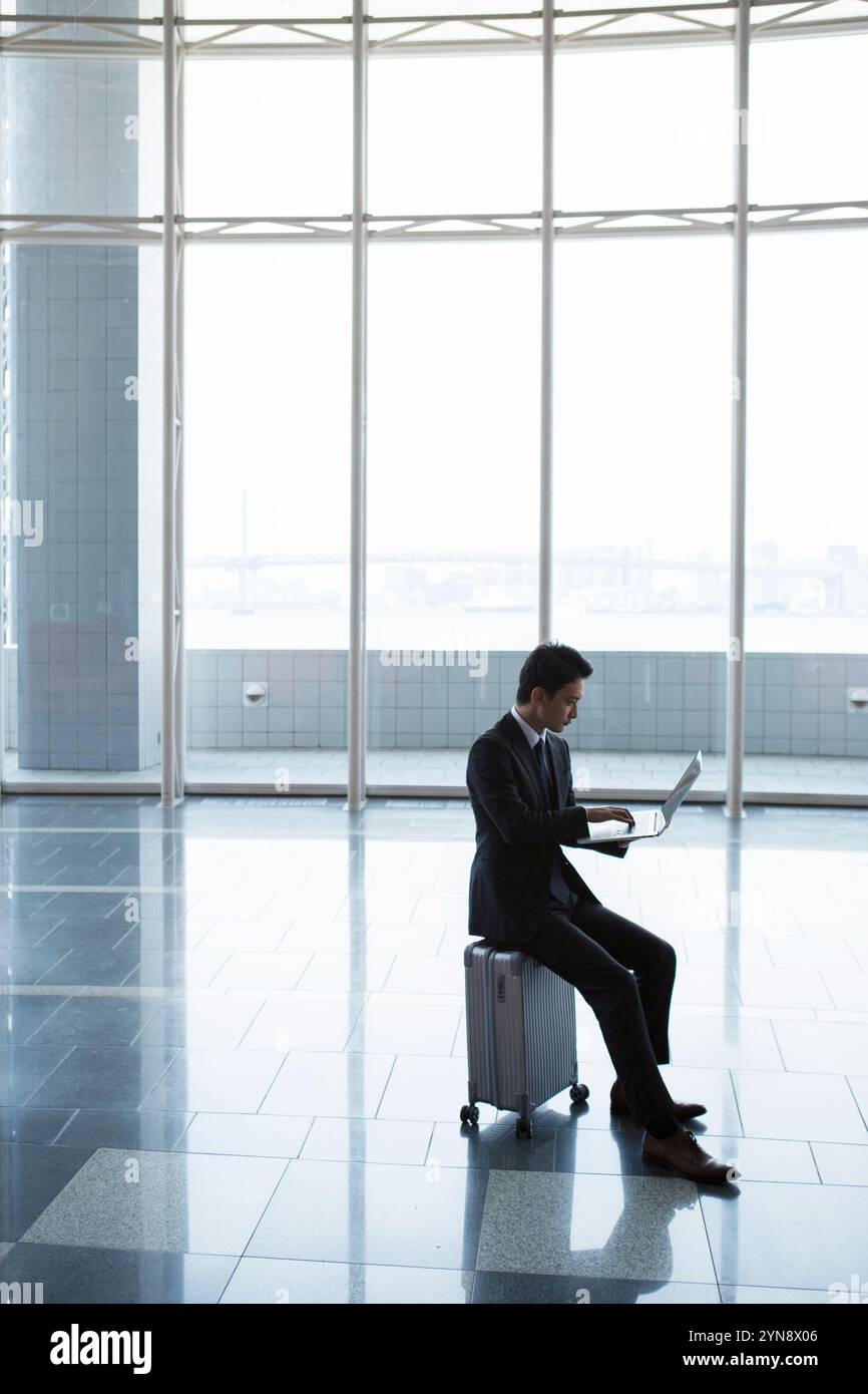 Man in his 20s in suit sitting on carrying case operating laptop ...