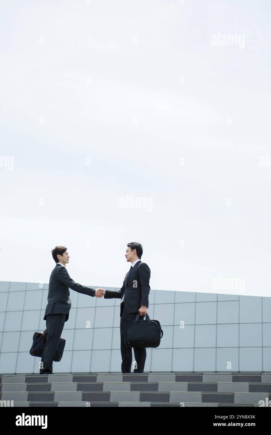 Two men in their 20s in suits shaking hands with their arms crossed ...