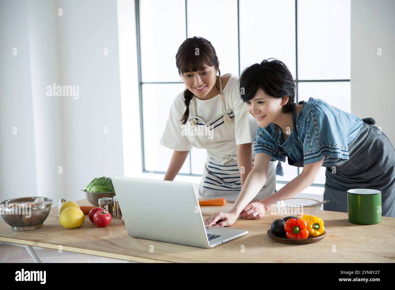 Two smiling women in their 20s cooking while checking their computers ...