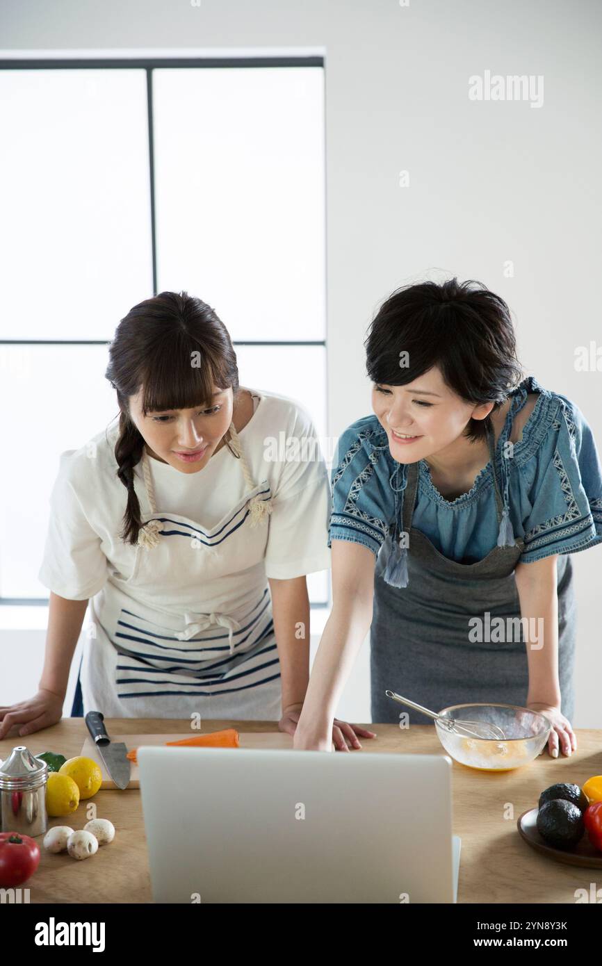 Two smiling women in their 20s cooking while checking their computers ...