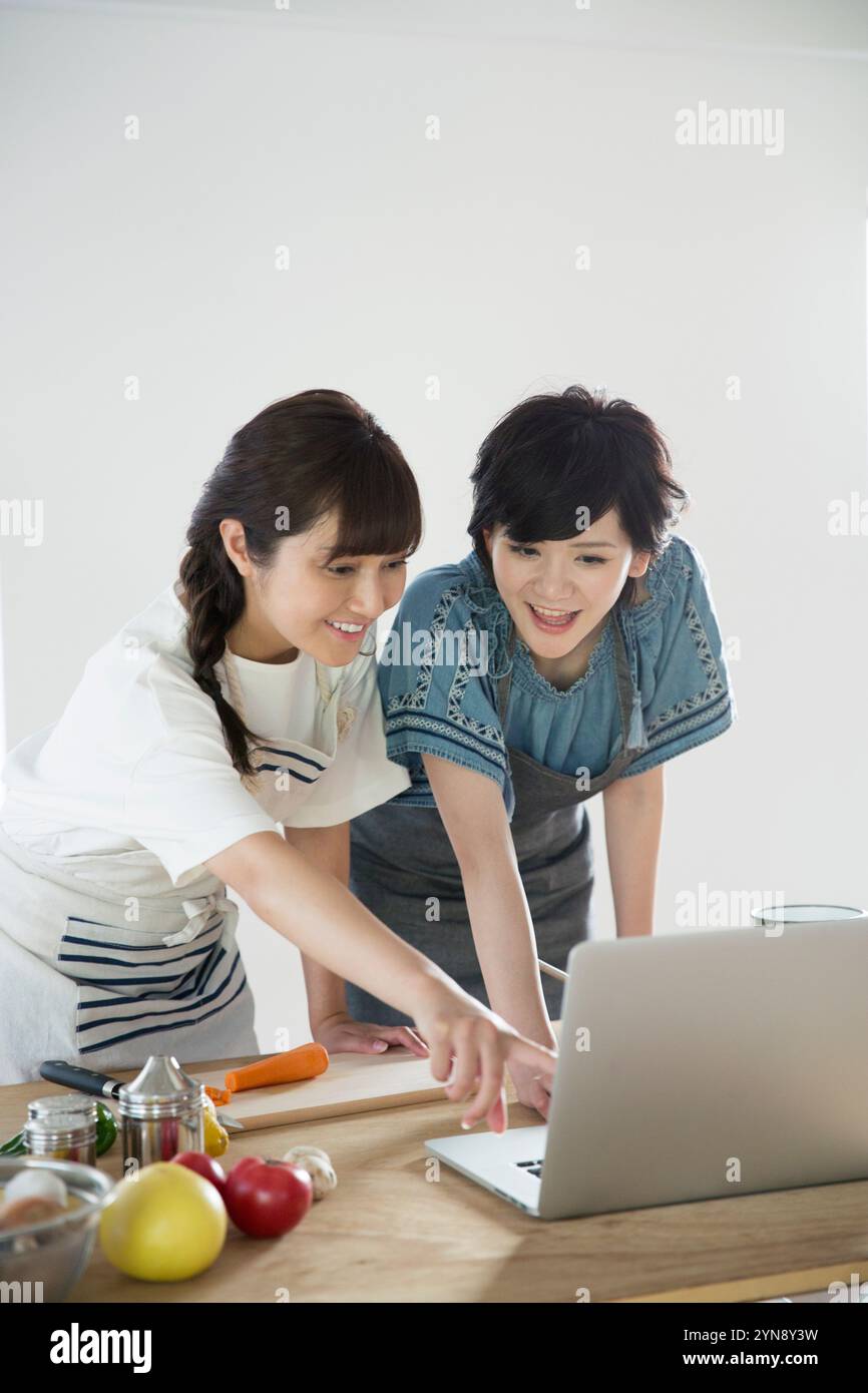 Two smiling women in their 20s cooking while checking their computers ...