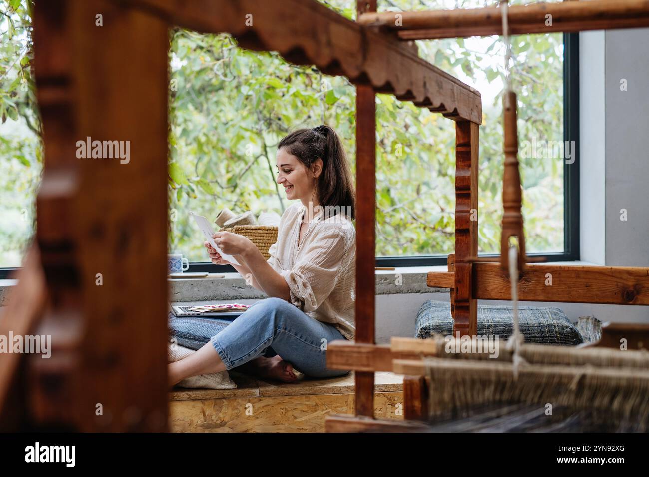 Young businesswoman looking at swatch book, sitting by wooden loom ...