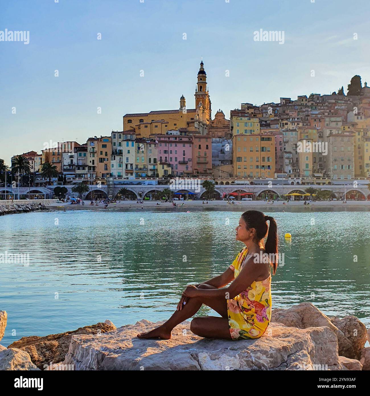 A young woman sits on a rock, gazing at the tranquil water reflecting ...