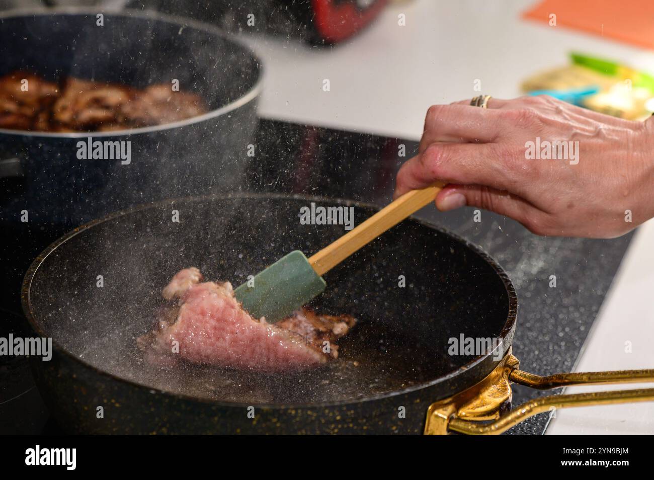 A hand skillfully flips sizzling meat in a hot pan, releasing aromatic ...