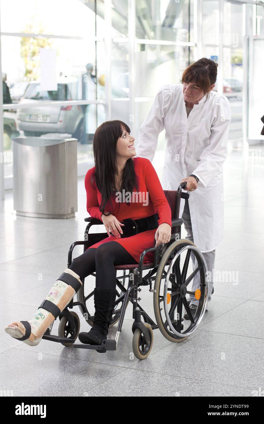 Young woman with leg in plaster sits in wheelchair, carer looks after ...