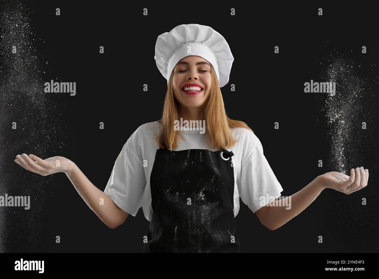Cheerful female chef playing with flying flour on black background ...