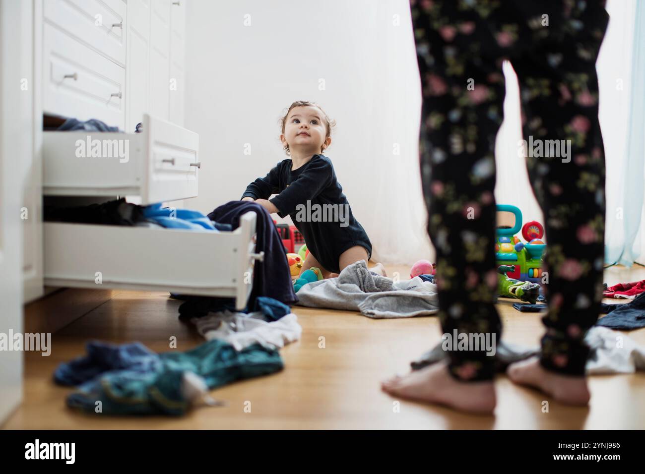 A young child is seated on the floor of a messy bedroom, surrounded by ...