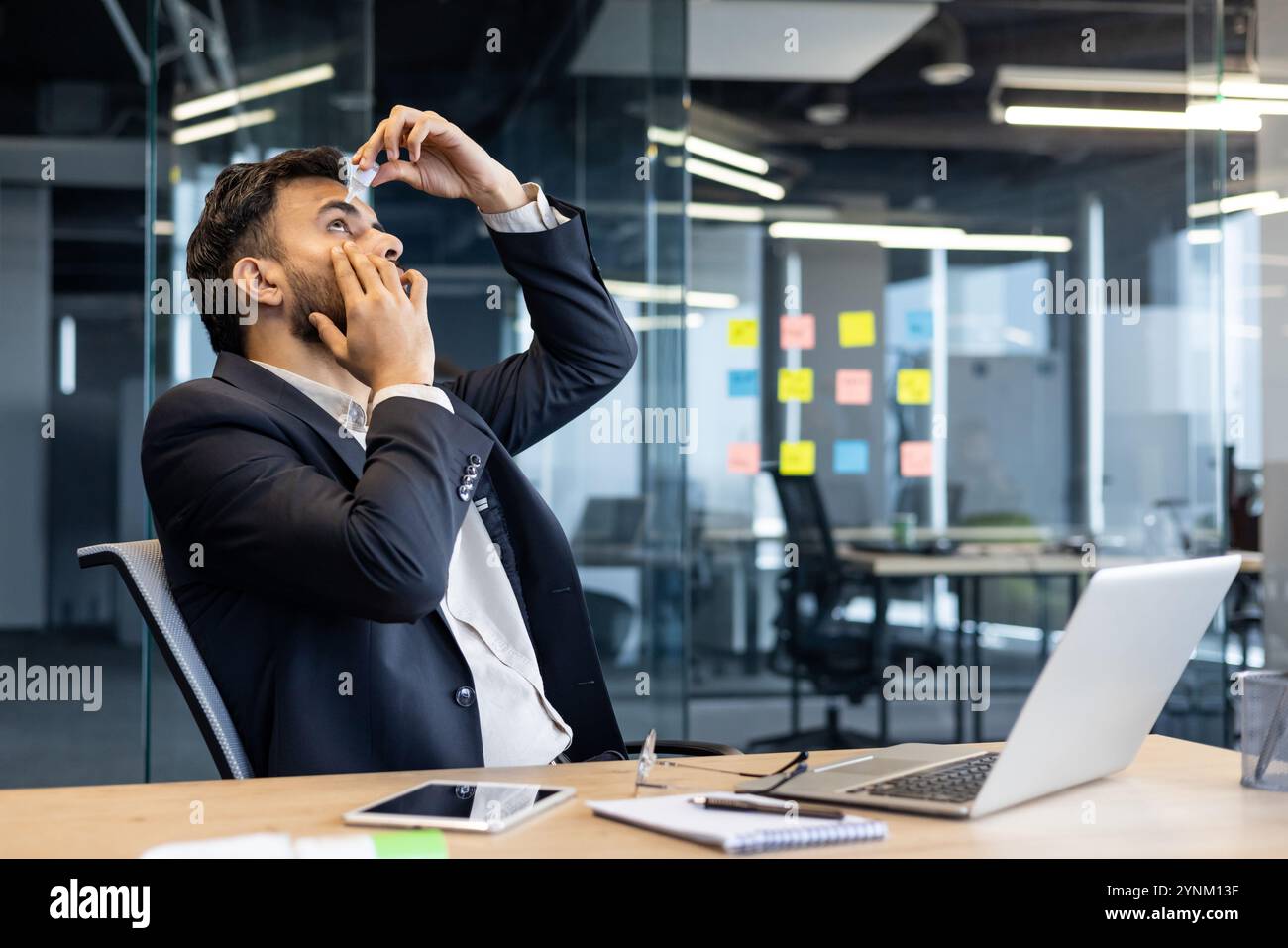 Office worker in suit applying eye drops at desk with laptop and phone ...