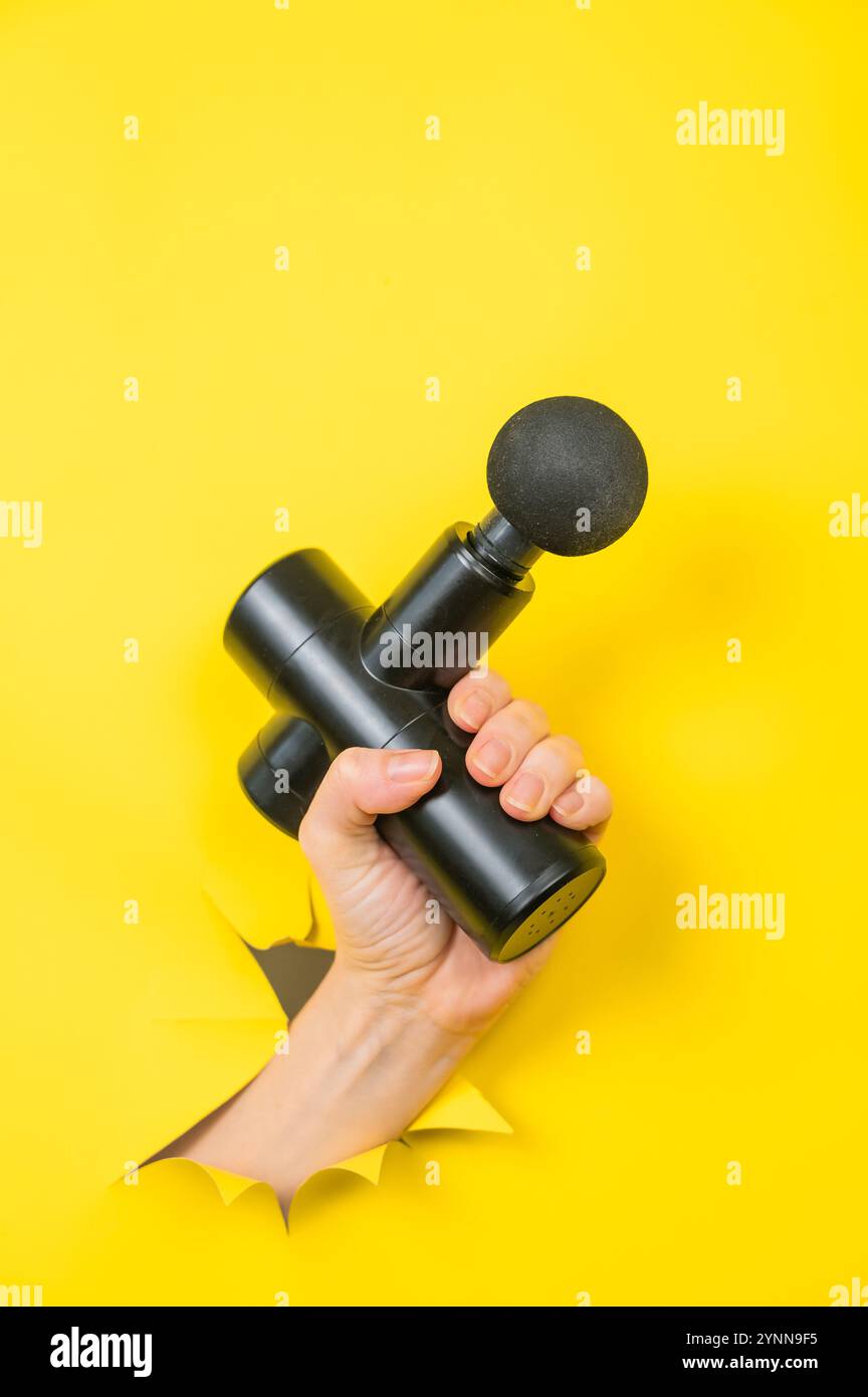 Female hand sticking out with massage gun from yellow paper background ...