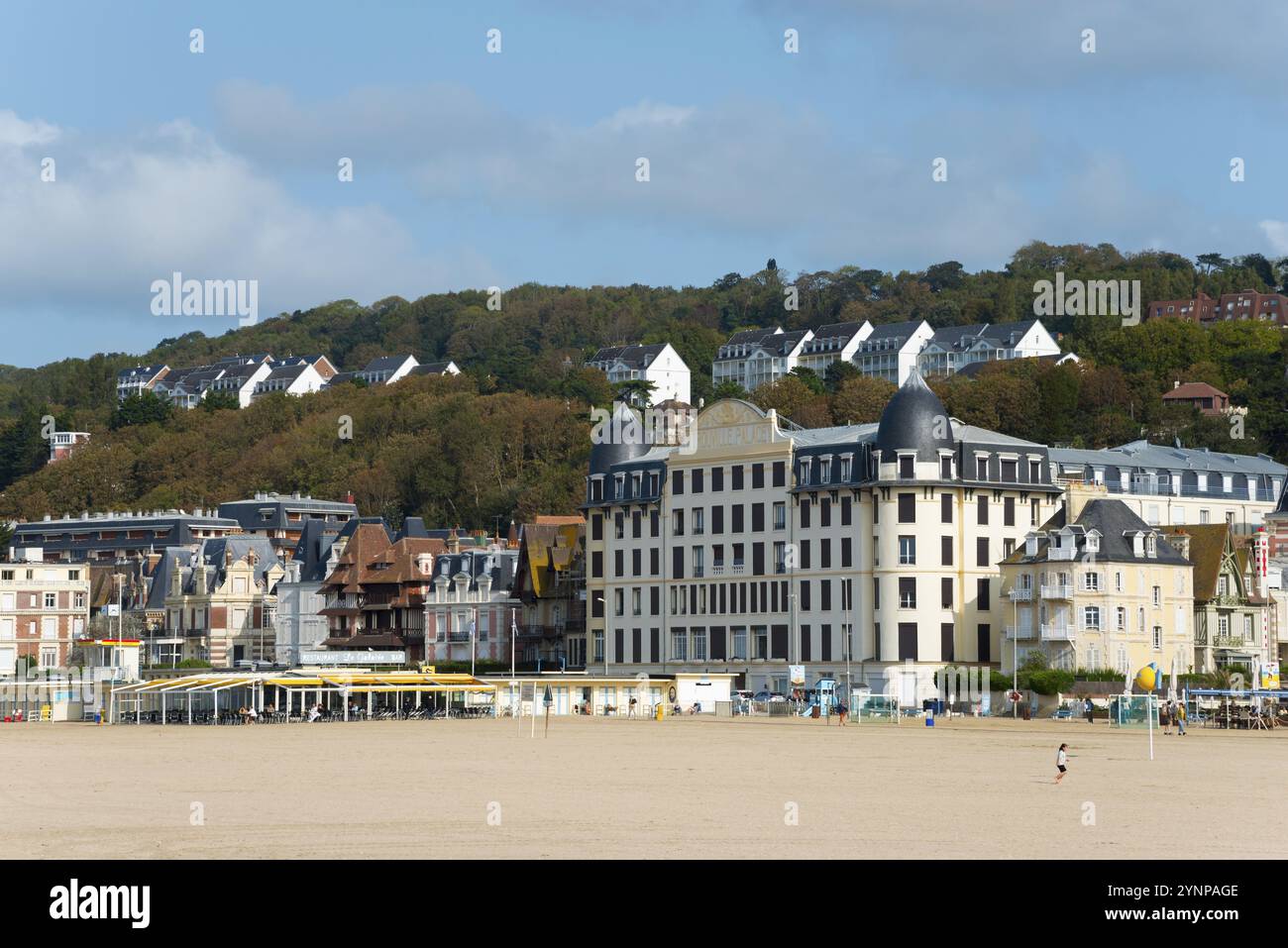 Modern buildings with striking towers on a wide beach in front of a wooded hilltop, on the right ...