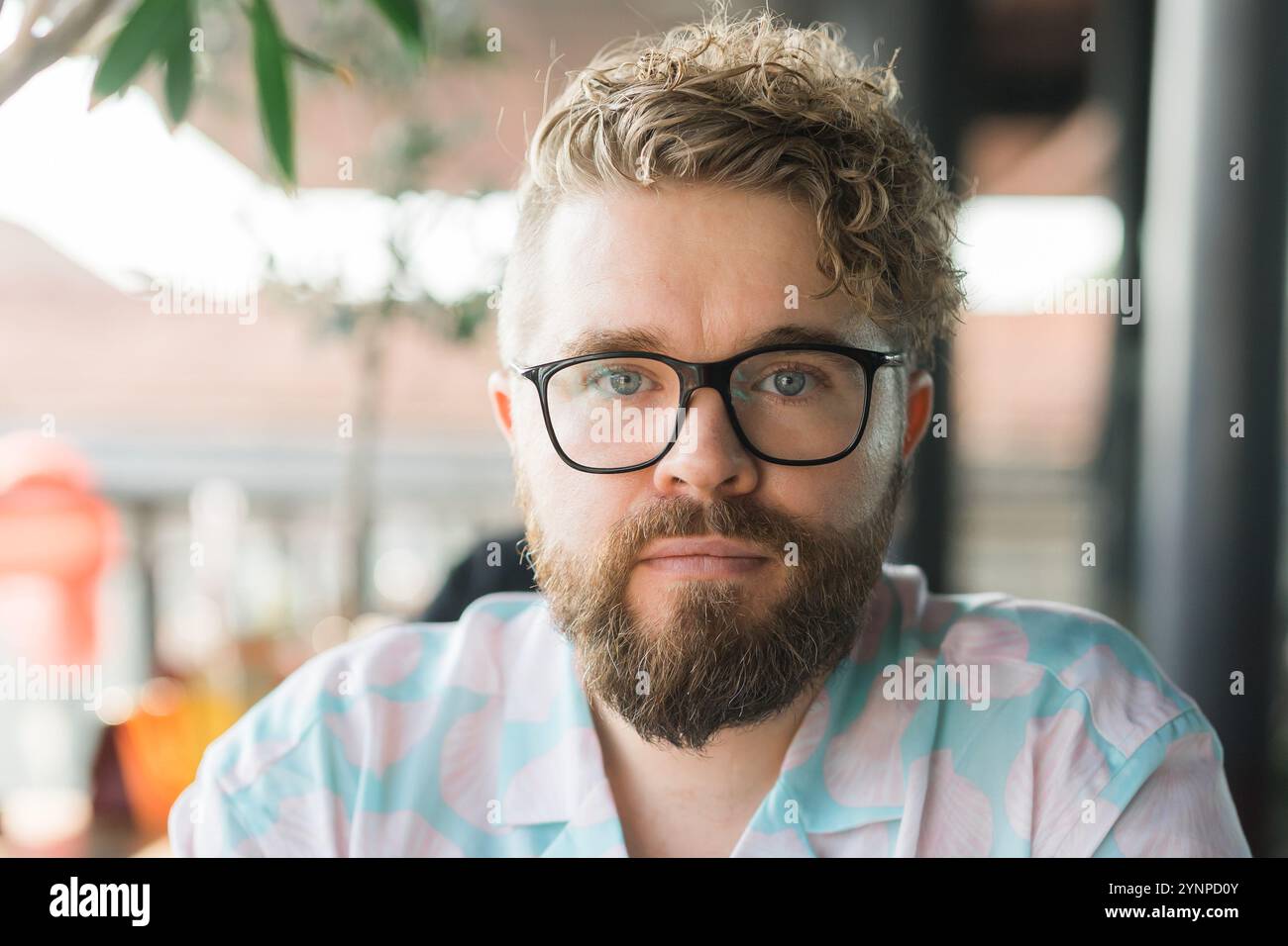 Close-up portrait of a man with glasses, curly hair, and beard, sitting outdoors at a cafe ...