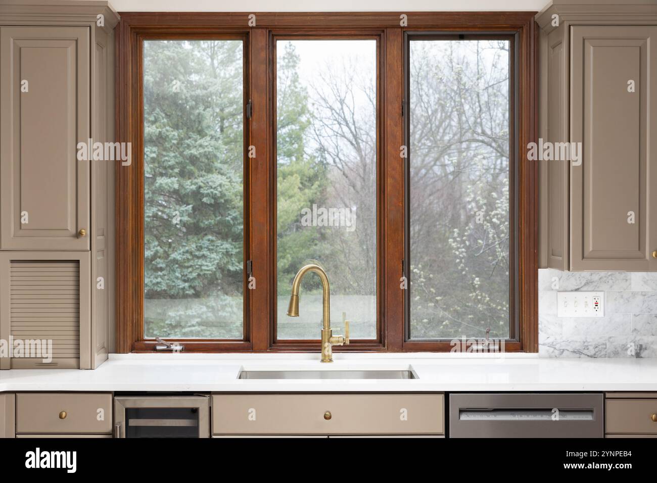 A gold kitchen faucet detail with brown cabinets, wood frame windows ...