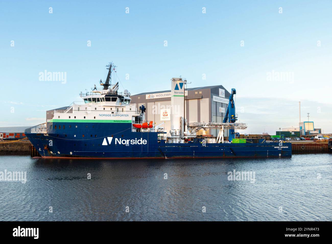 A large blue offshore supply vessel docked at a bustling shipyard under ...