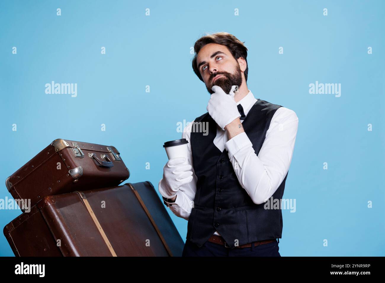 Thoughtful doorman drinks coffee in studio, standing next to luggage ...