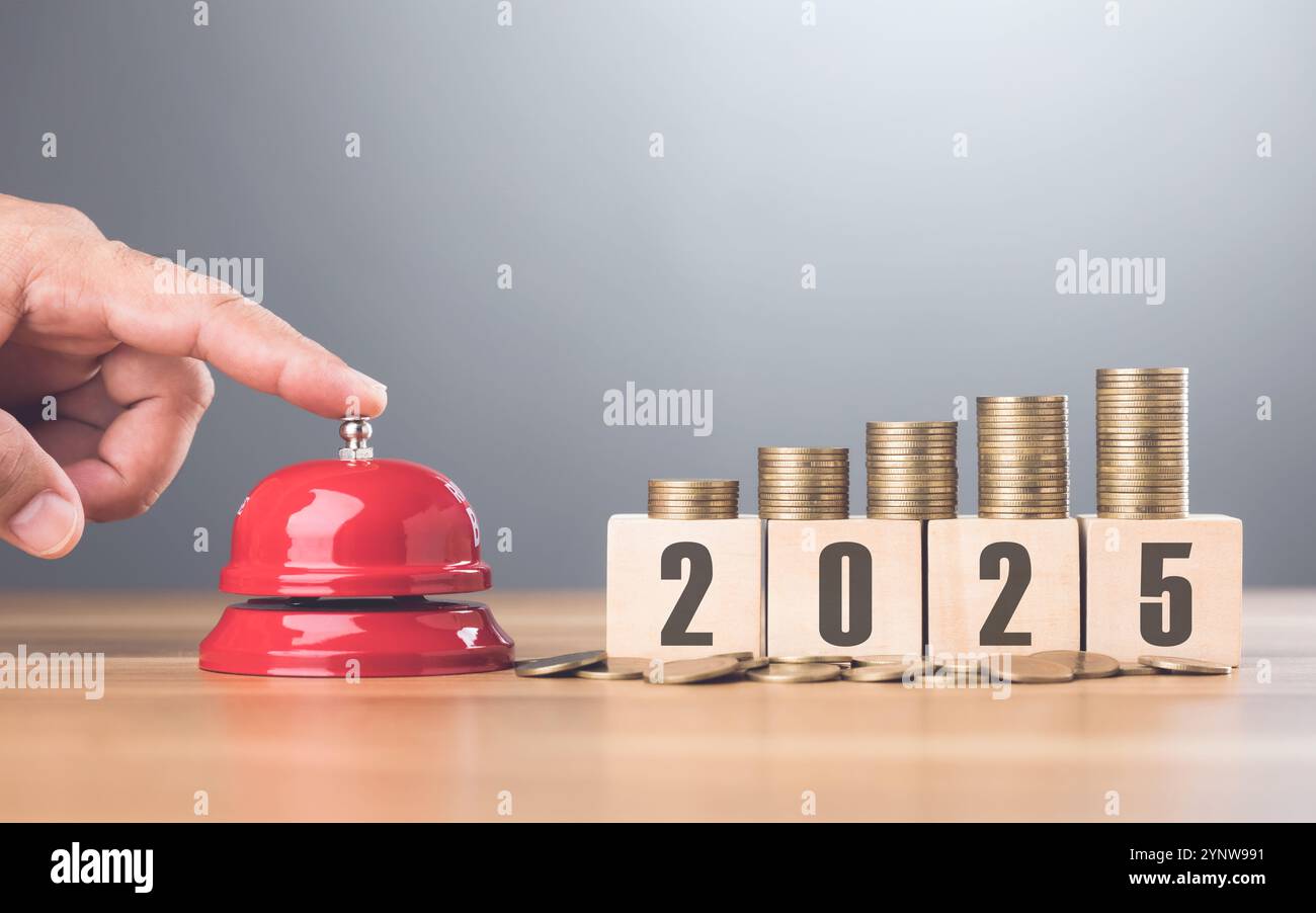 A hand presses a red bell next to a stack of coins on a wooden block ...