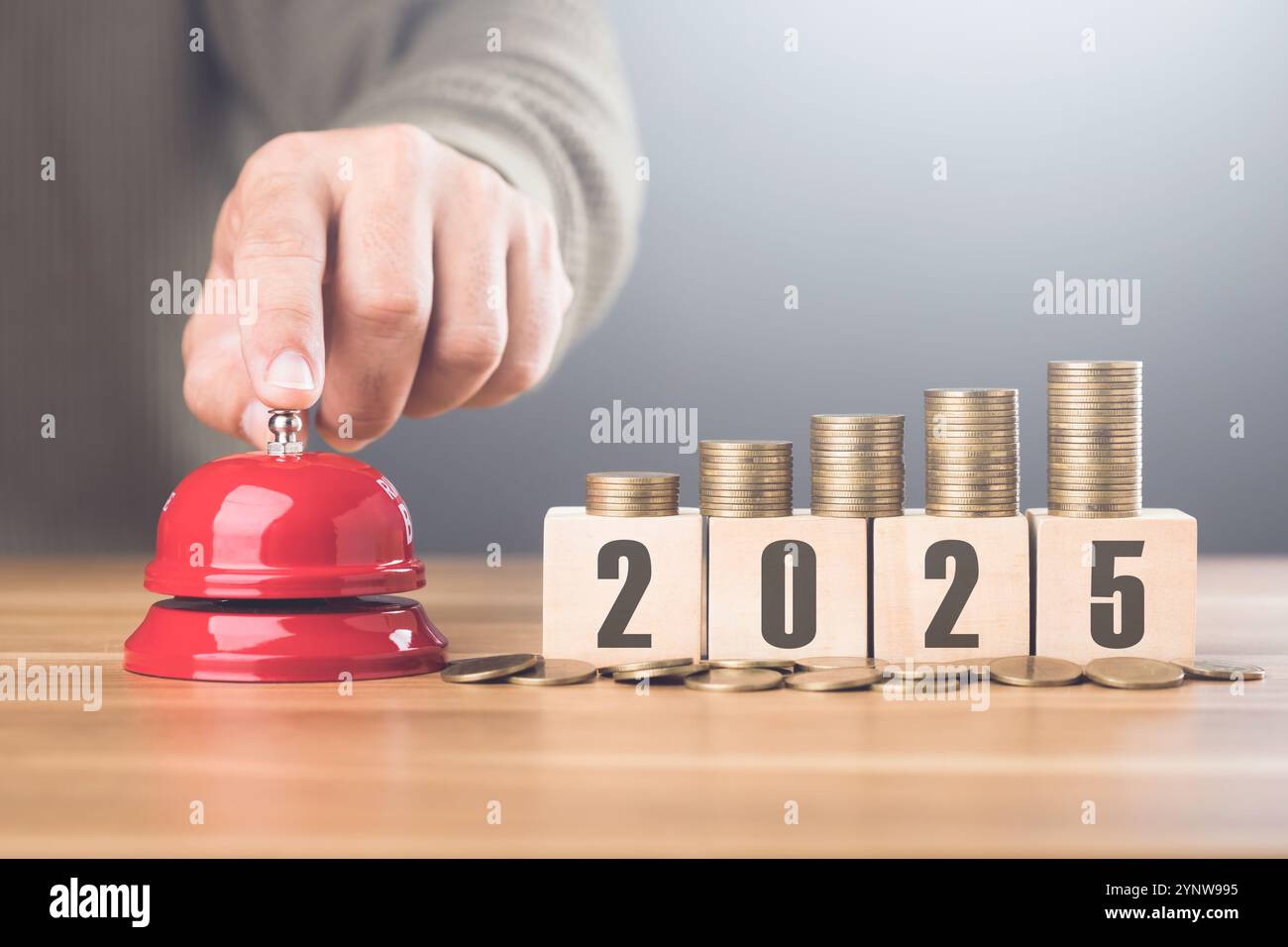 A hand presses a red bell next to a stack of coins on a wooden block ...