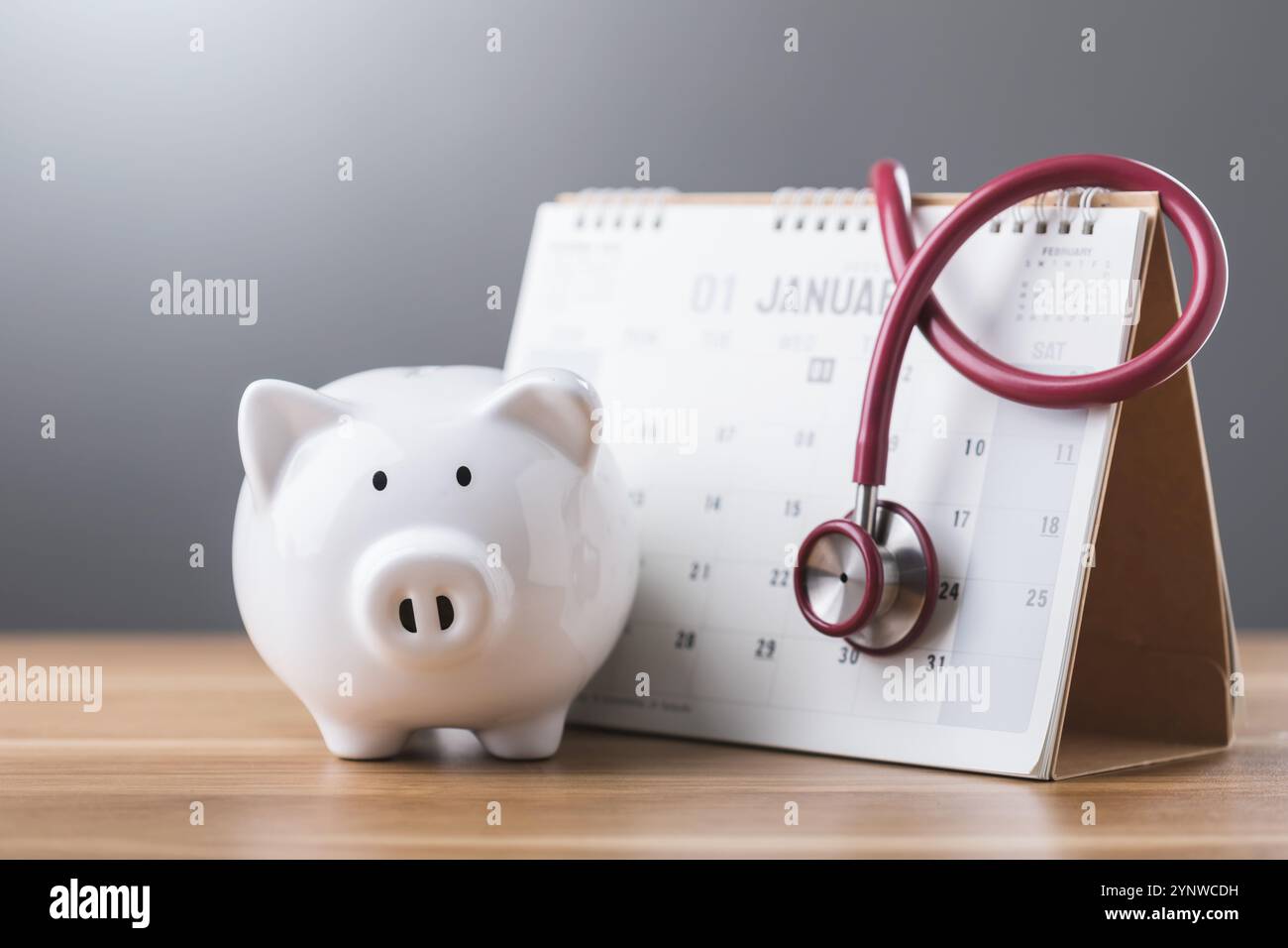 A white piggy bank, a calendar, and a stethoscope on a table symbolize ...