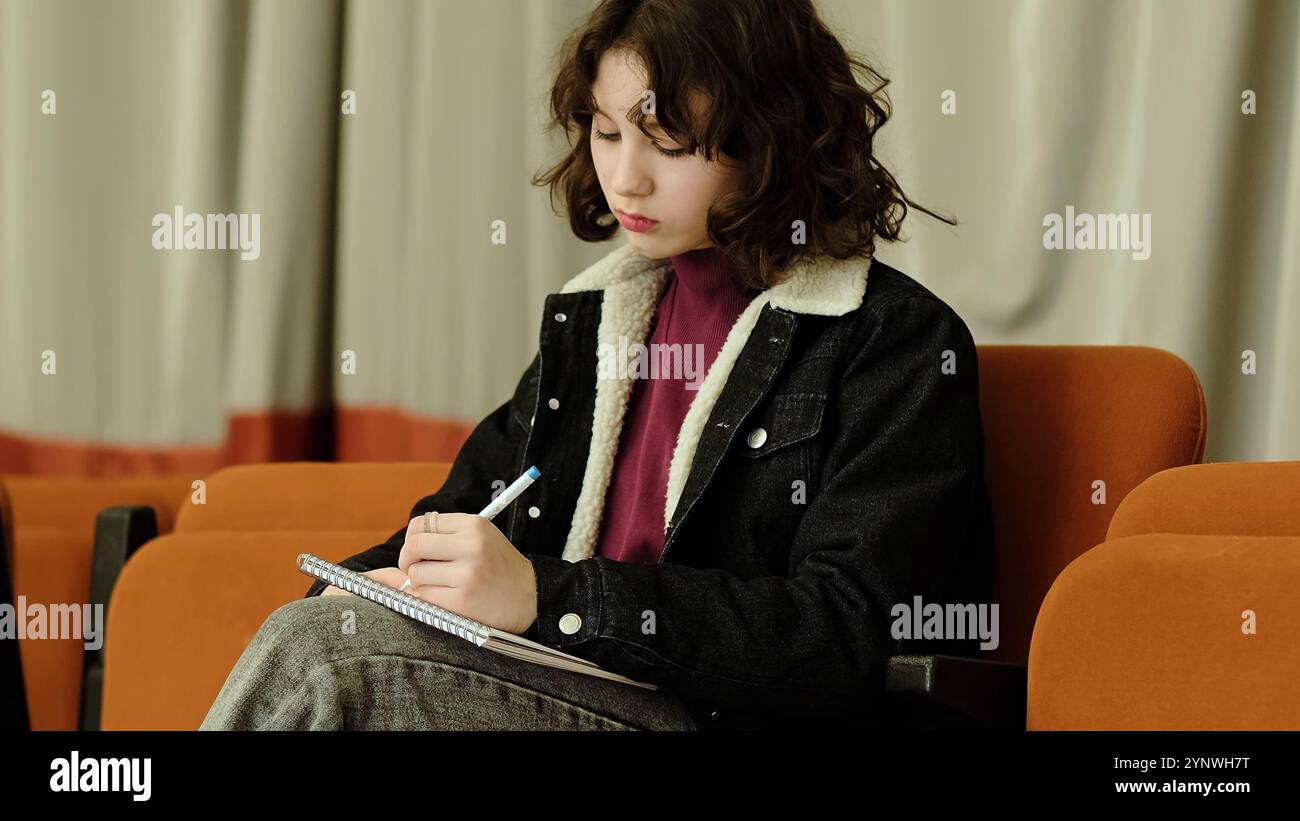 A focused girl sits in an auditorium chair, immersed in her notebook ...