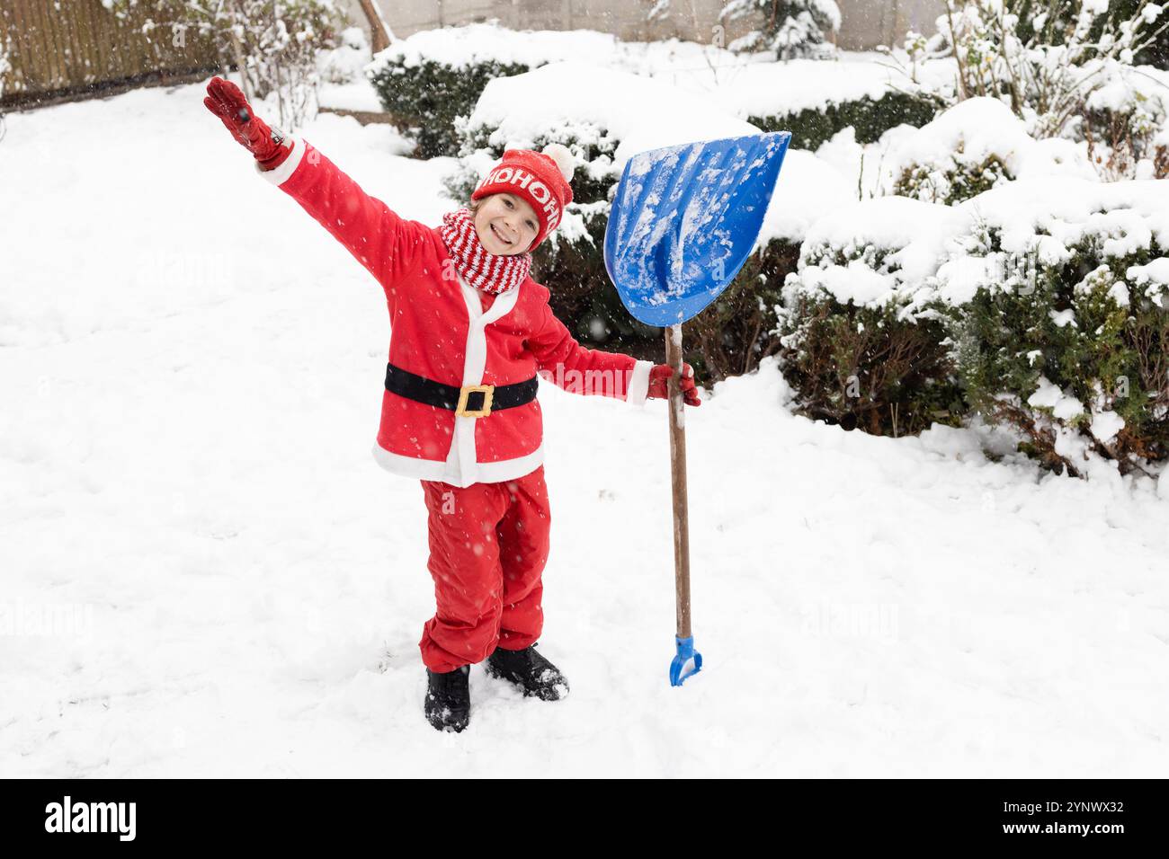 portrait of a cheerful boy in a red Santa suit with a large snow shovel ...