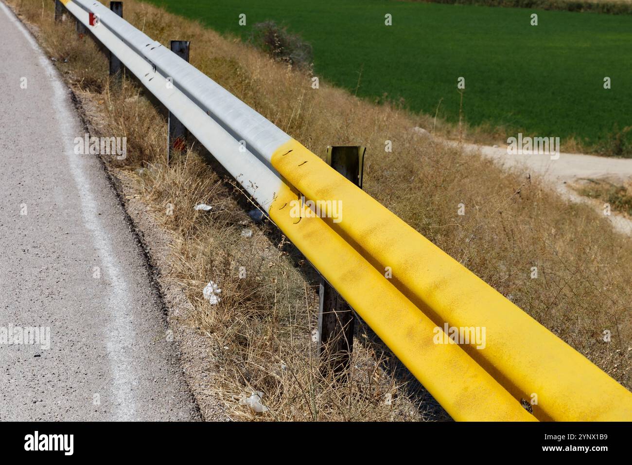 A rugged yellow safety barrier runs alongside a dry road, with lush ...