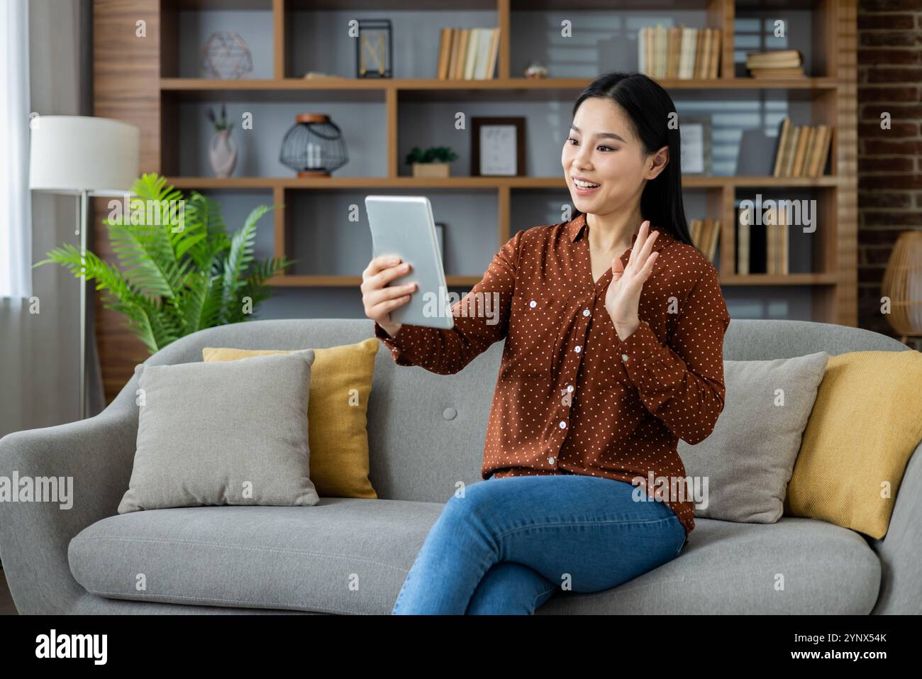 Asian woman smiling, sitting on sofa using tablet for virtual ...