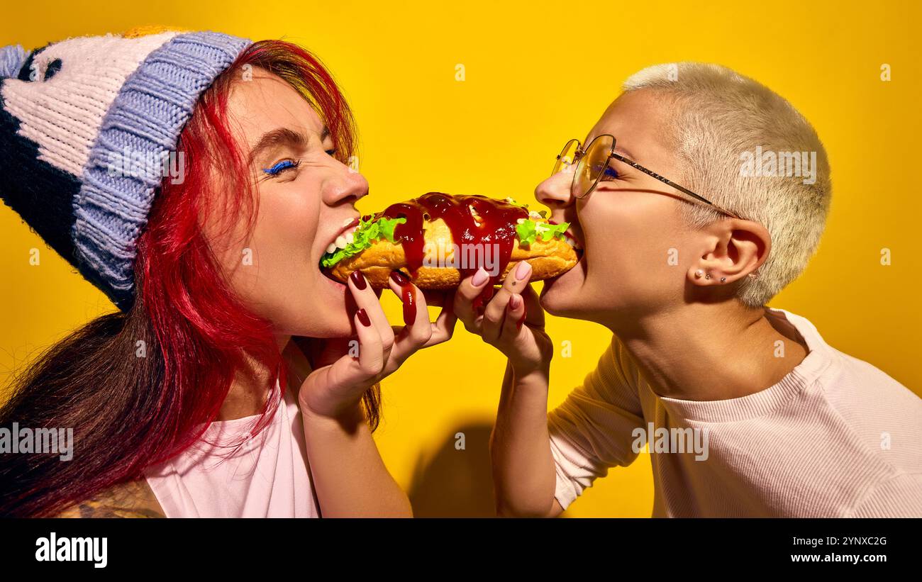 Two women emotional and passionately eating hot dog, biting it from two ...