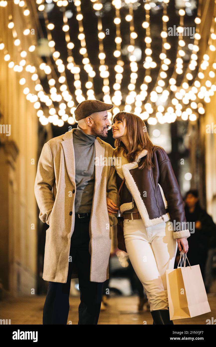 A joyful couple carrying shopping bags, strolling under twinkling ...