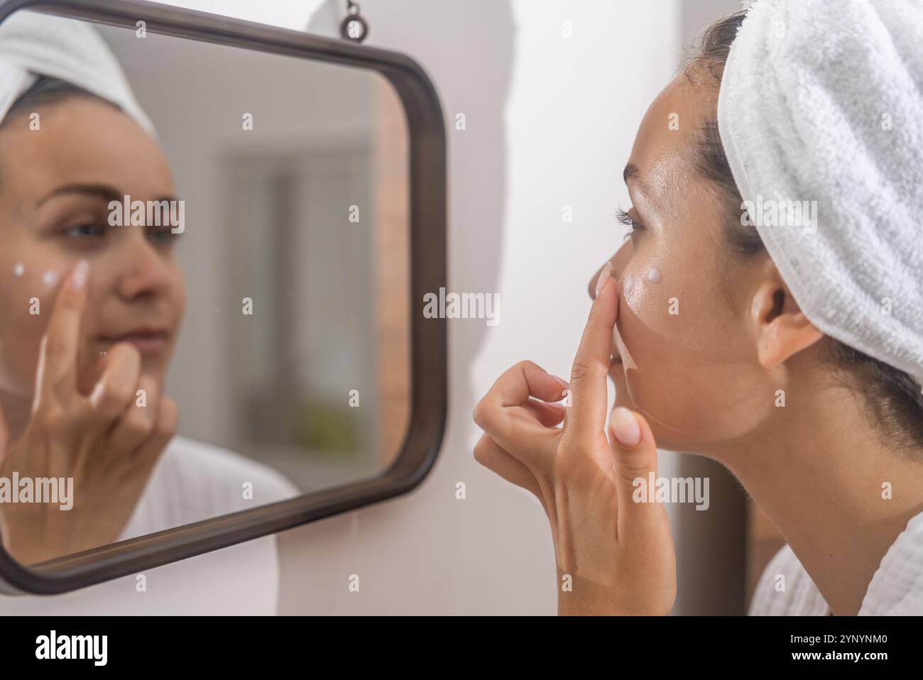 Happy woman applying moisturizing cream under eye looking in mirror in ...