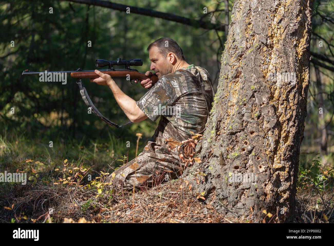 Hunter in camouflage clothing leaning against a tree, aiming a rifle ...