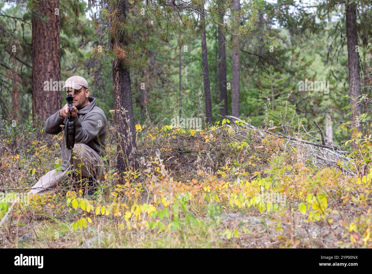 A hunter in a kneeling position aims his rifle at game, peering through ...