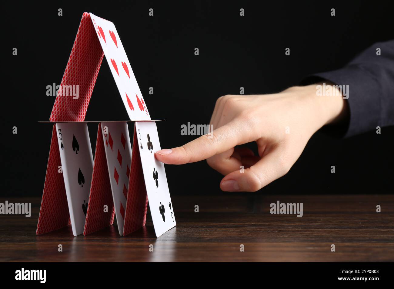 Woman destroying house of playing cards at wooden table against black ...