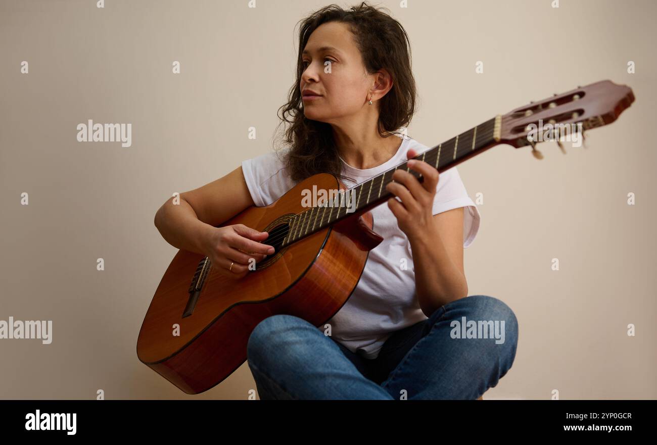 A woman sits cross-legged while playing an acoustic guitar, creating a ...