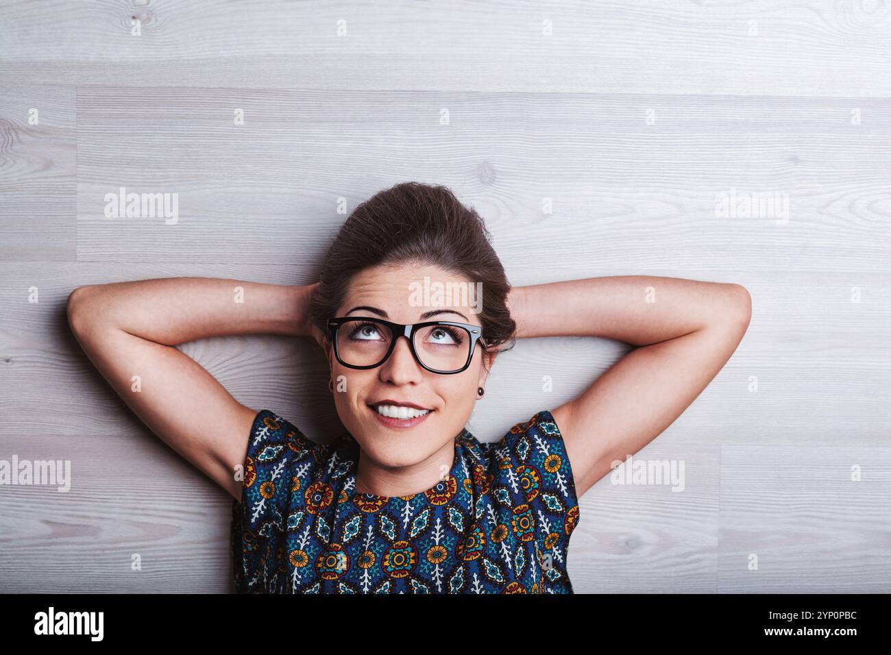 Young woman with eyeglasses is lying on a wooden floor with her hands ...