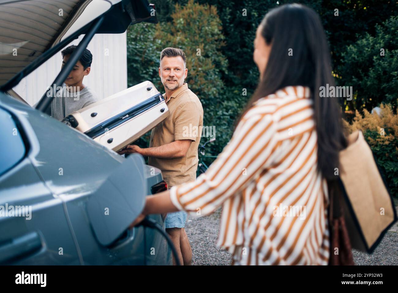 Rear view of woman charging car while man loading luggage with son in ...